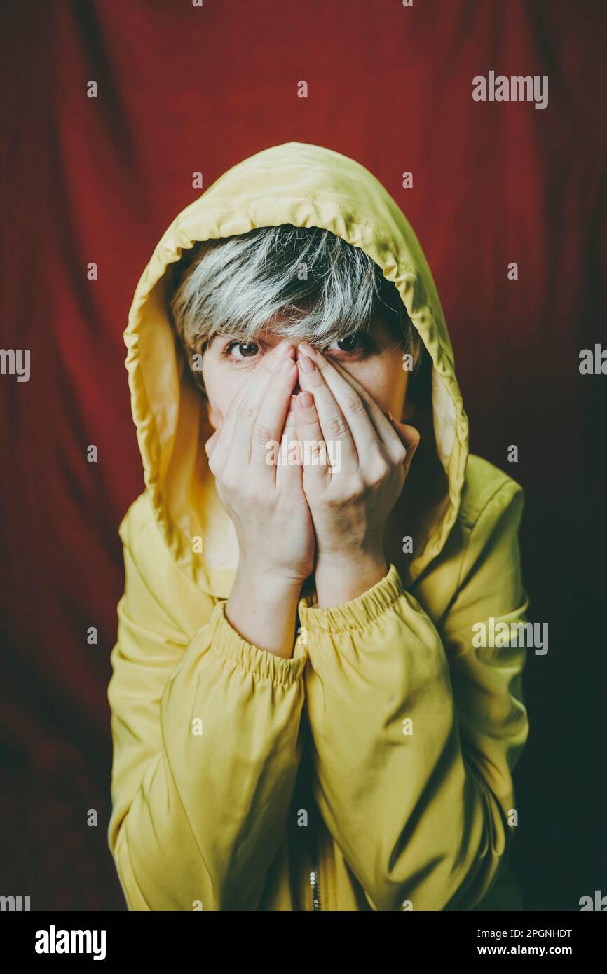 Woman wearing yellow hooded jacket covering mouth against red backdrop ...
