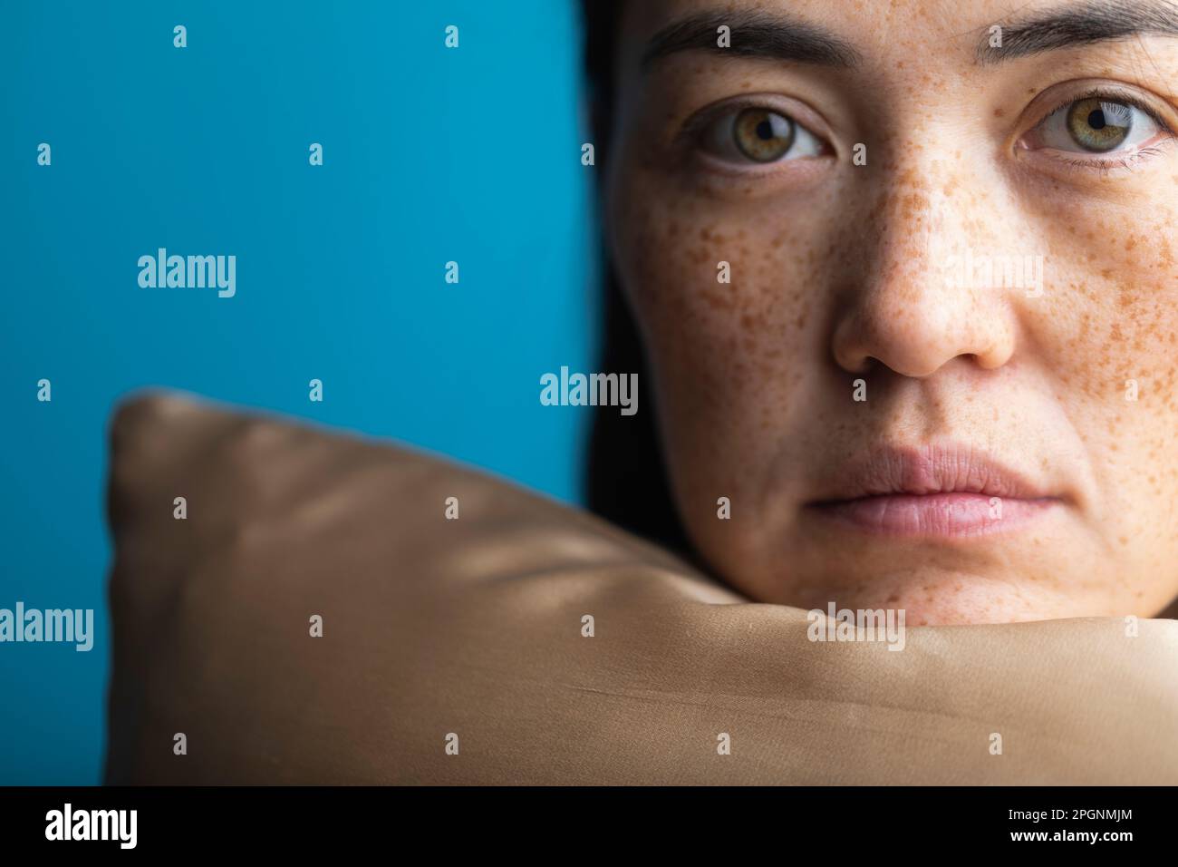 Woman with freckles resting chin on silk pillow against blue background ...