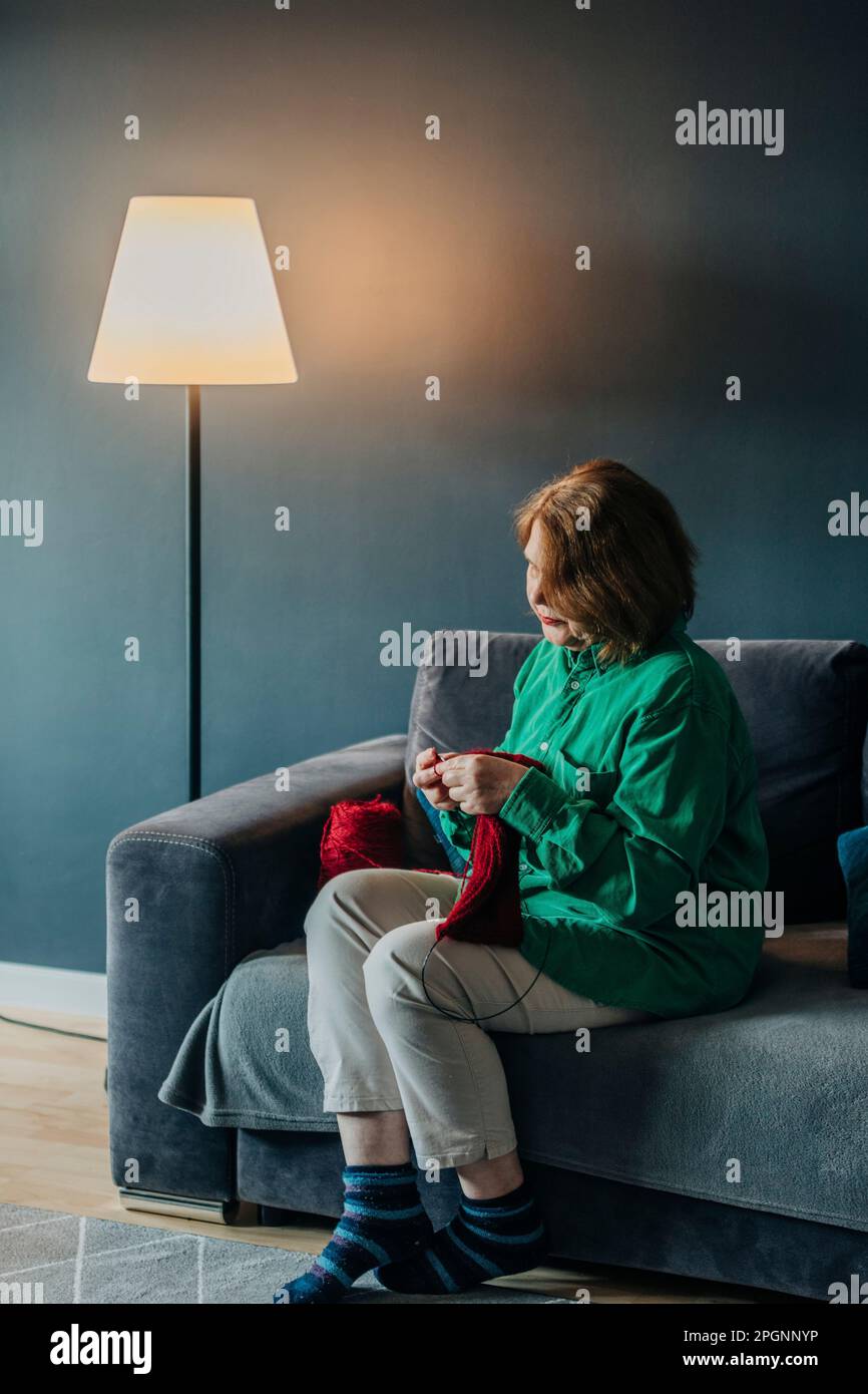 Senior Woman Knitting Sweater Sitting On Sofa By Illuminated Lamp At