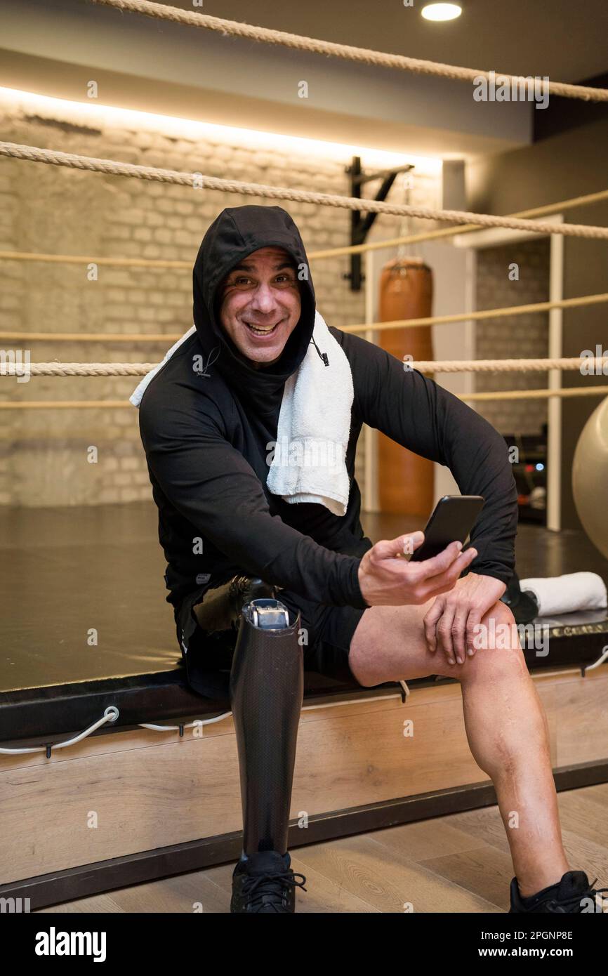 Happy man with disability sitting on boxing ring holding smart phone in ...