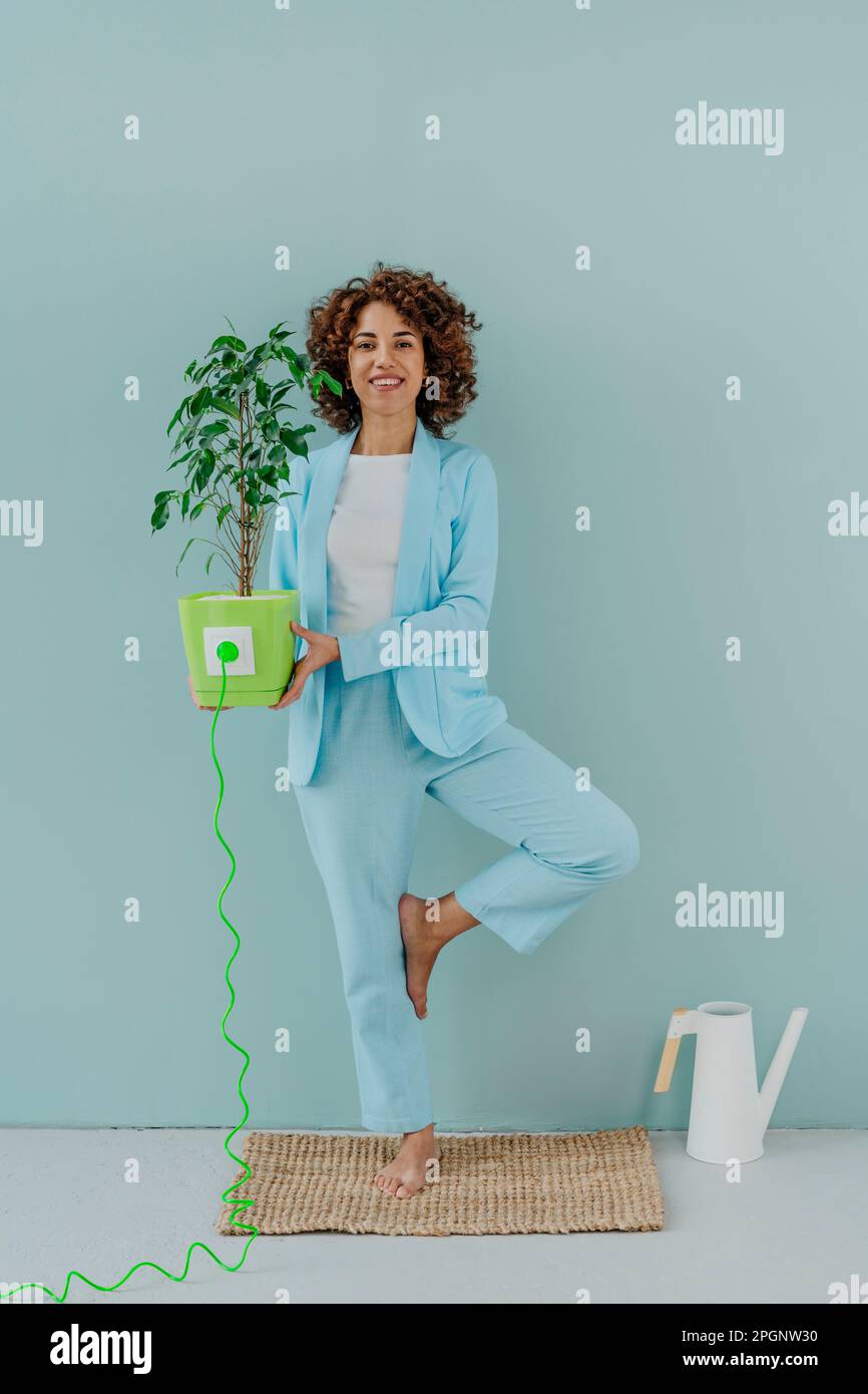 Happy woman practicing tree pose with potted plant in front of blue ...