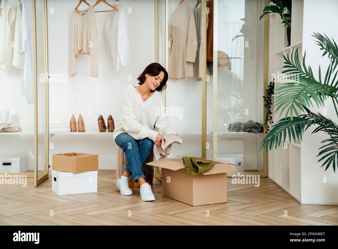 Young woman putting clothes in box sitting in front of closet at home ...