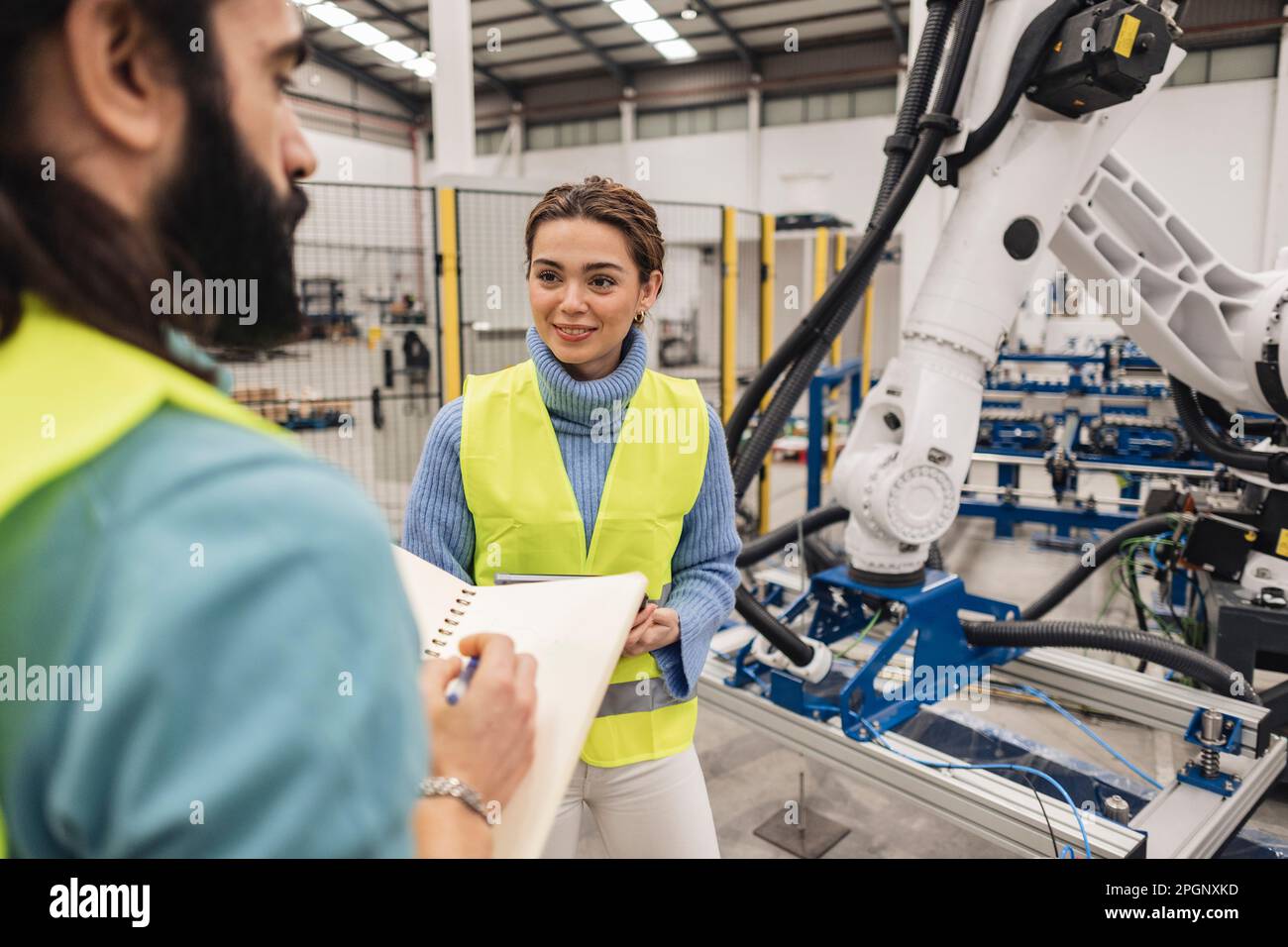 Smiling engineer looking at colleague with note pad standing by robotic ...