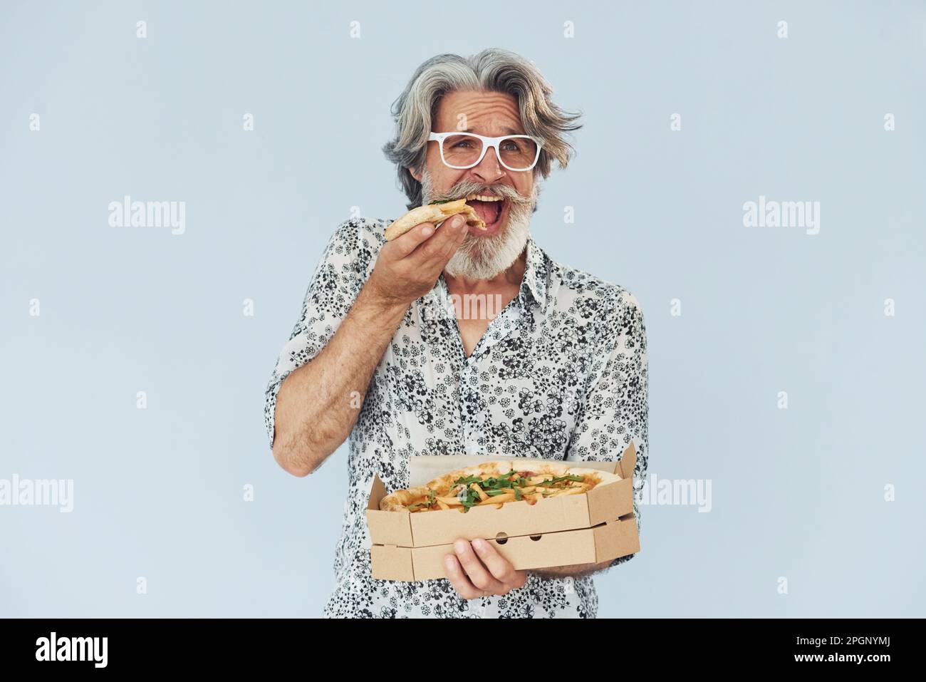 Senior stylish modern man with grey hair and beard indoors eats pizza ...