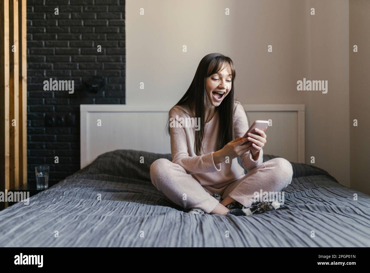 Surprised young woman using mobile phone sitting cross-legged on bed in bedroom Stock Photo - Alamy