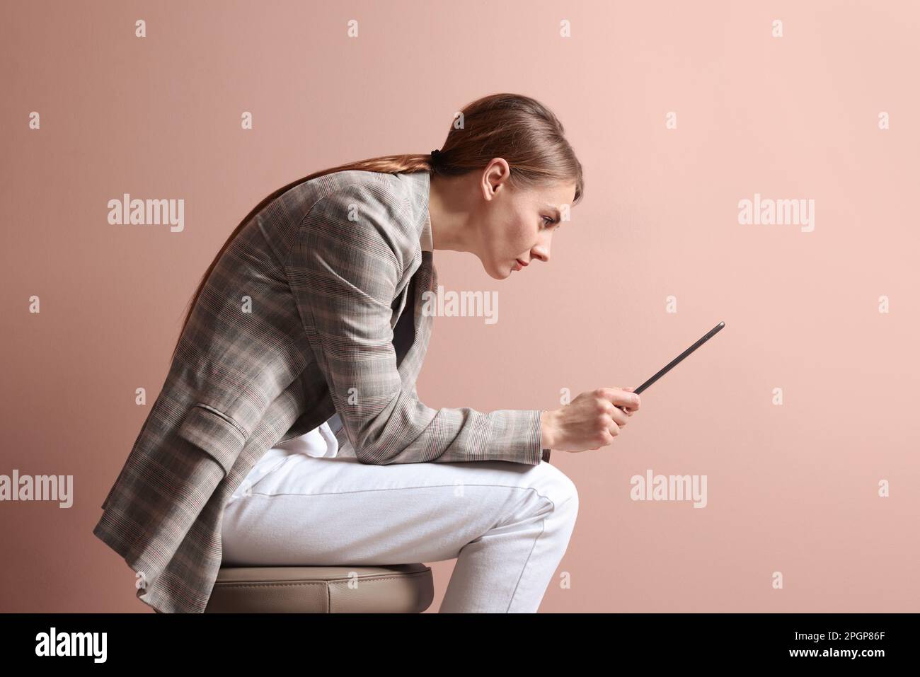 Woman with bad posture using tablet while sitting on stool against pale ...