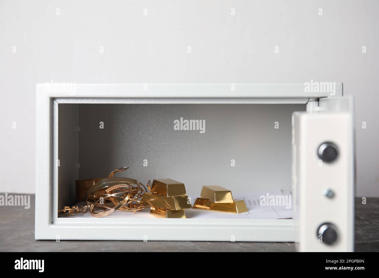 Open steel safe with gold bars and jewelry on grey table against light ...