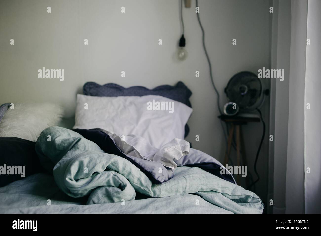 An image of an unmade bed in a bedroom with a fan in the corner Stock ...