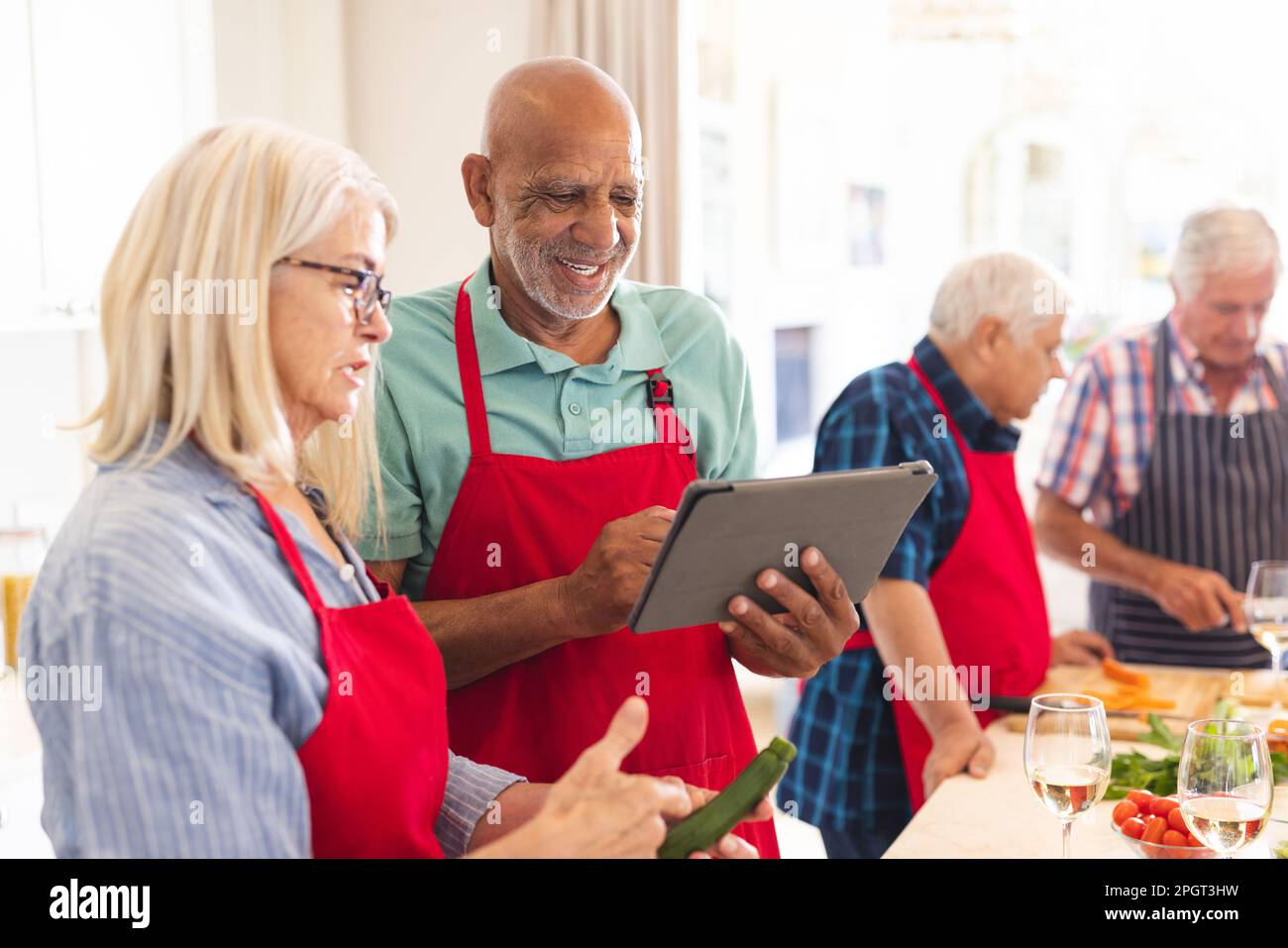 Happy group of diverse senior friends in aprons, using tablet during ...