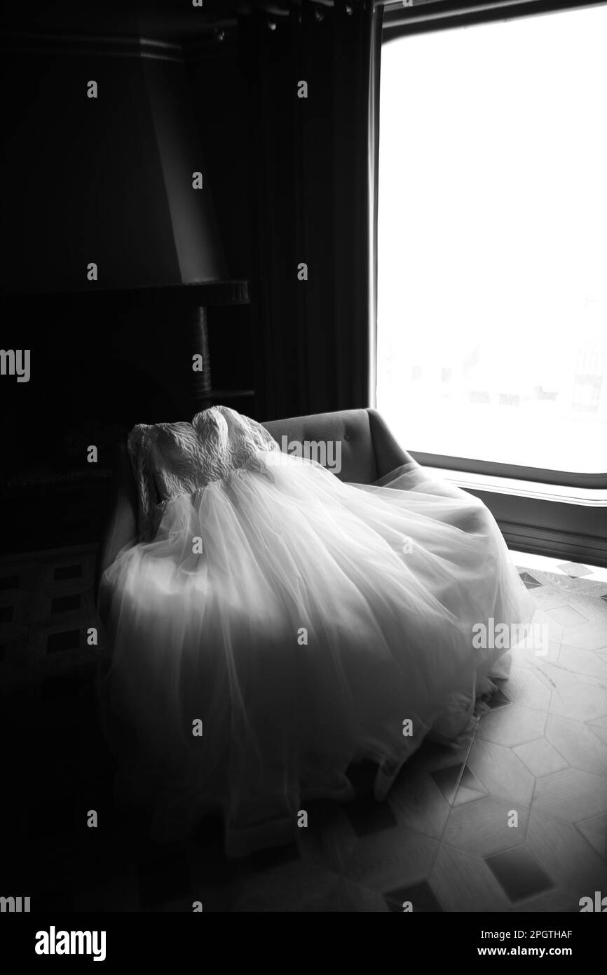 A greyscale of a bridal gown draped over a chair in front of a sunlit ...