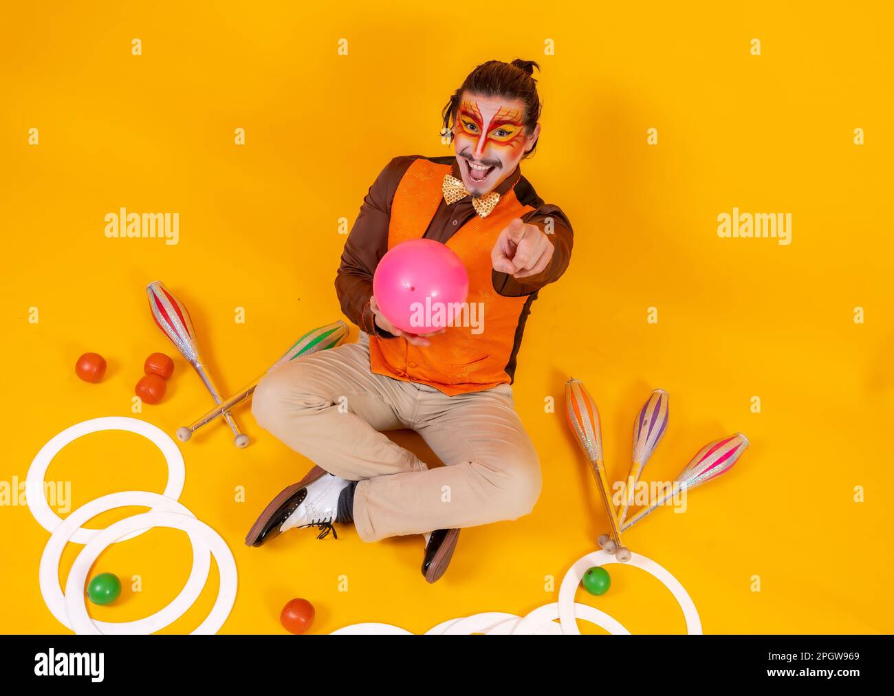 A cheerful circus performer sitting on the ground surrounded by various ...
