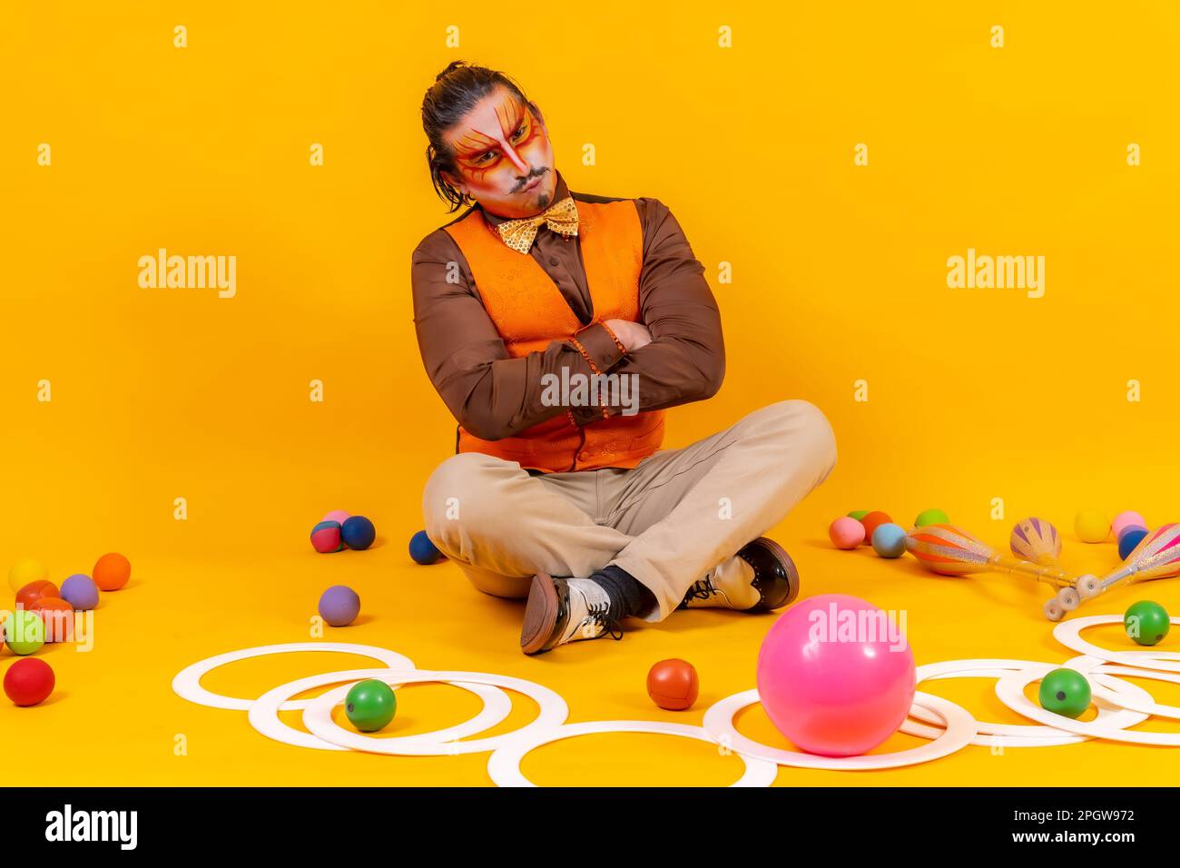 A cheerful circus performer sitting on the ground surrounded by various ...