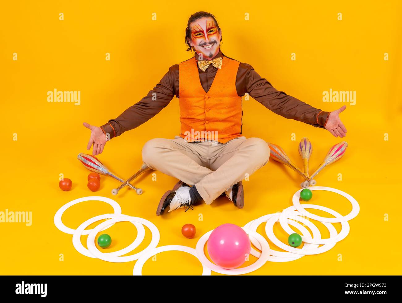 A cheerful circus performer sitting on the ground surrounded by various ...