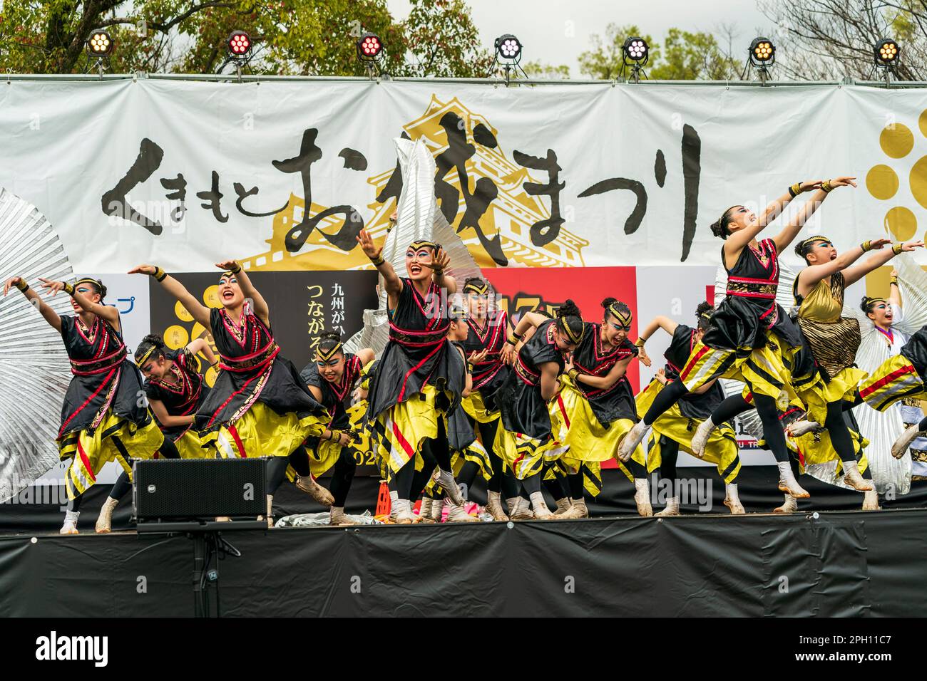 Group of about 25 young Japanese women in yellow and black costumes on an outdoor stage ...