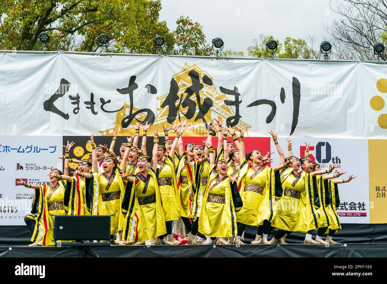 Group of about 25 young Japanese women in yellow costumes on an outdoor stage performing Yosakoi ...