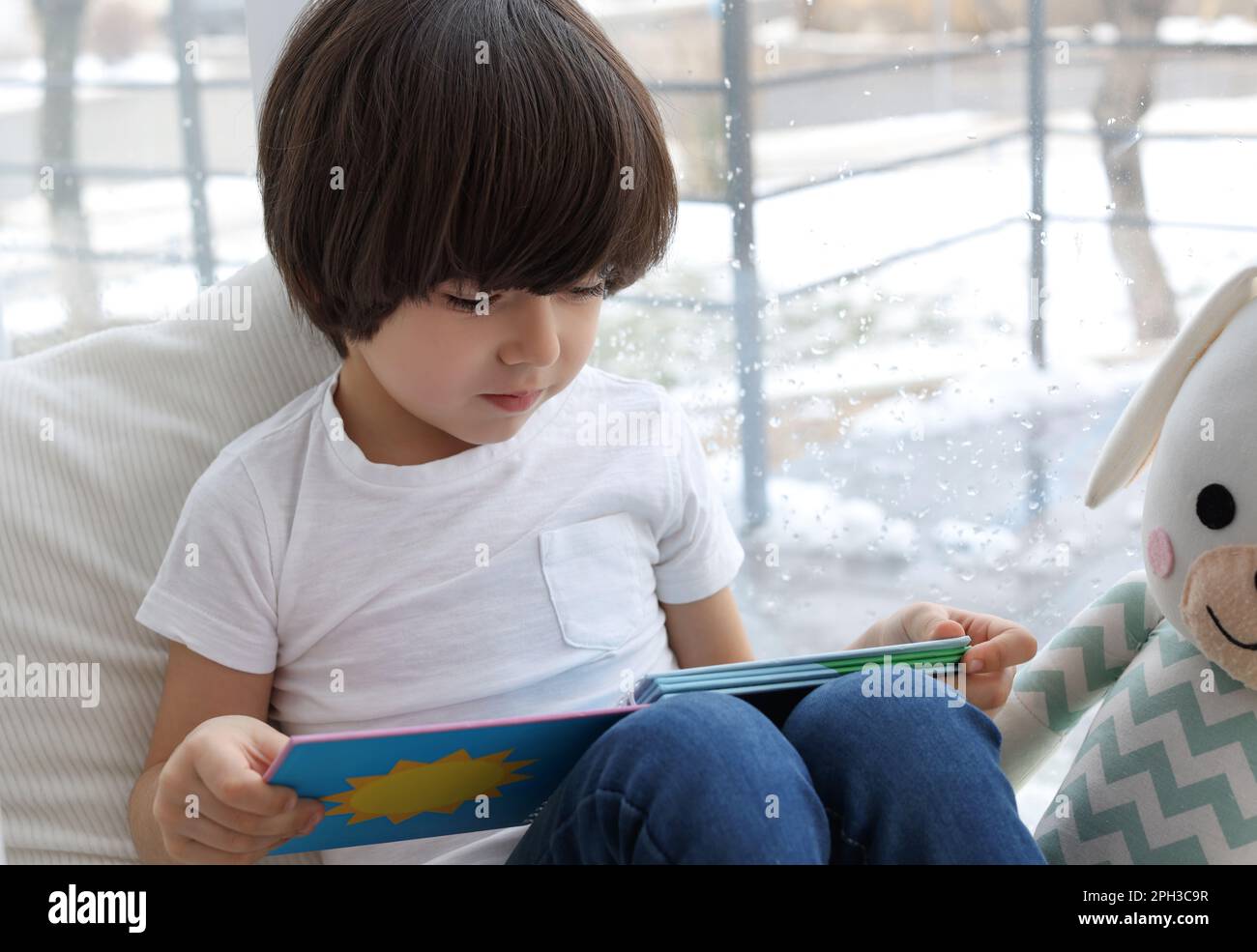 Cute little boy with toy reading book near window at home Stock Photo ...
