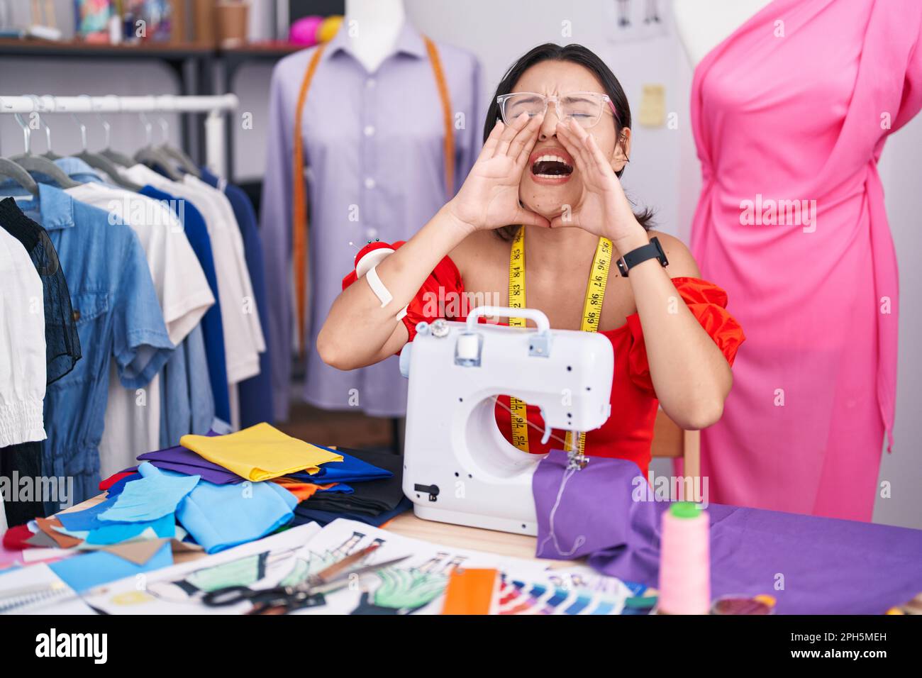 Hispanic young woman dressmaker designer using sewing machine shouting ...