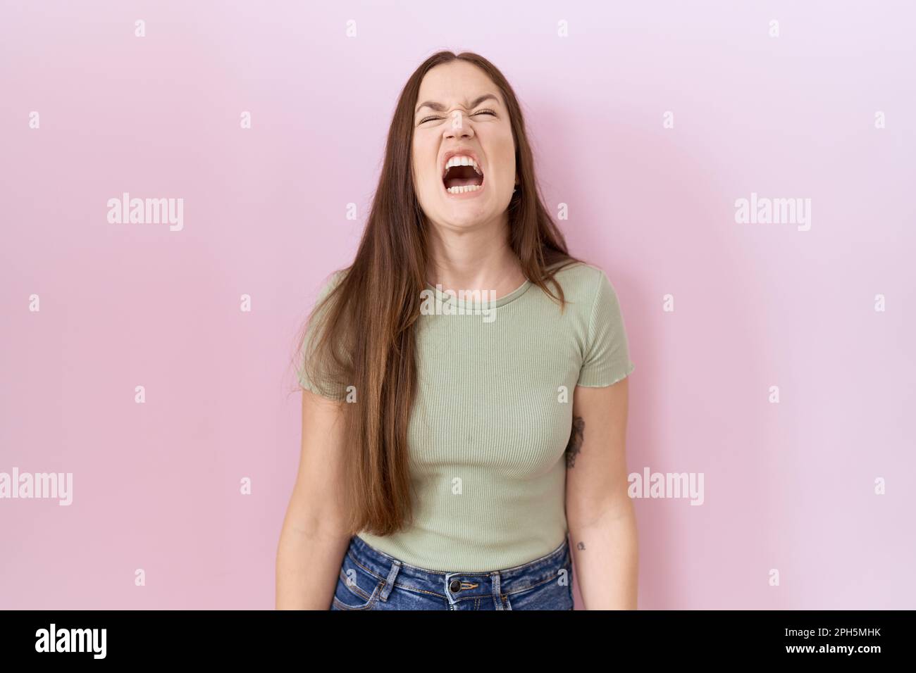 Beautiful brunette woman standing over pink background angry and mad ...