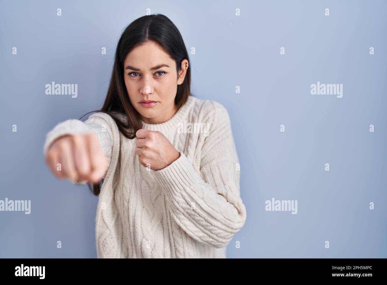 Young brunette woman standing over blue background punching fist to ...