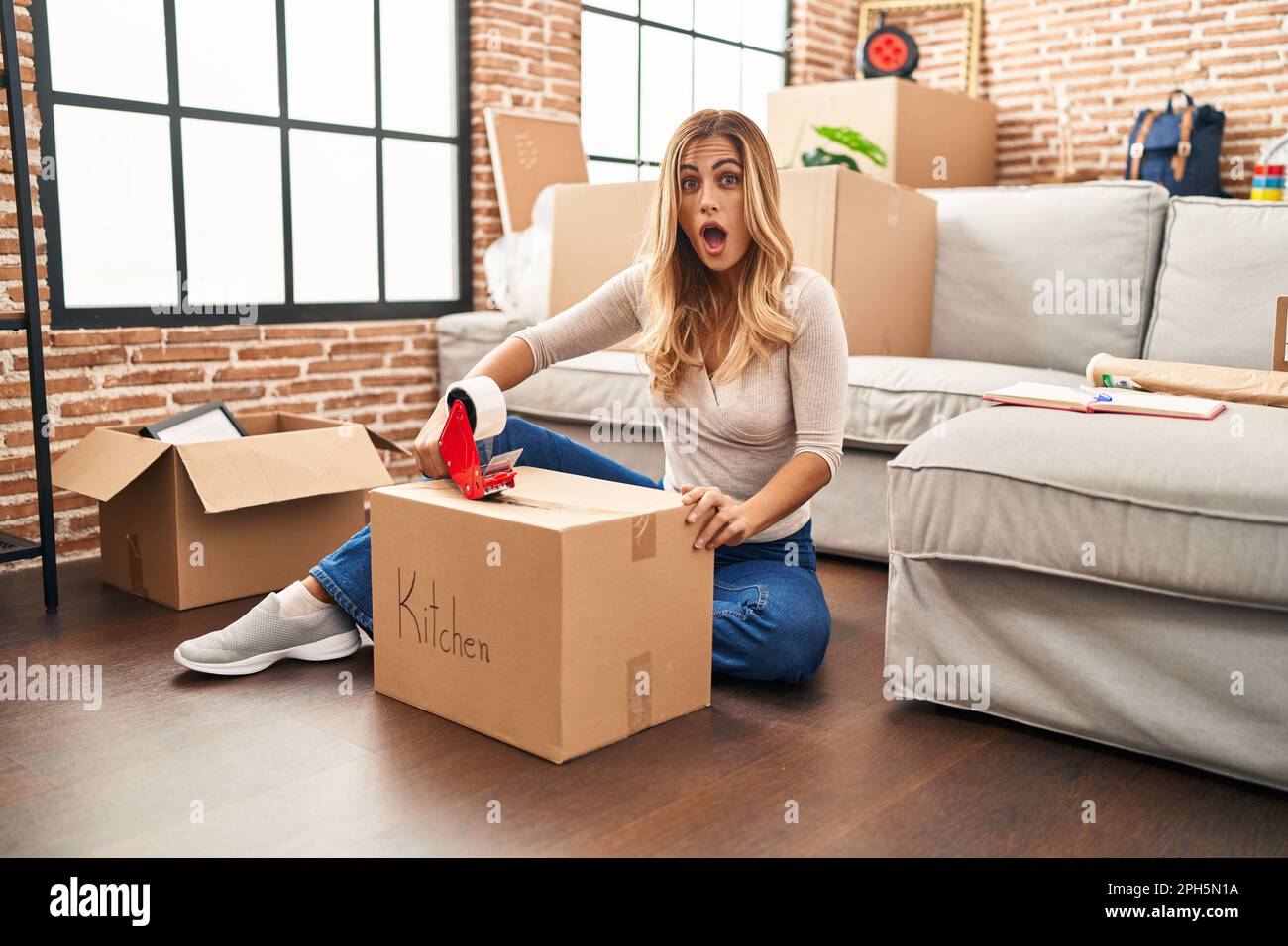 Young blonde woman sitting on the floor closing boxes home scared and ...