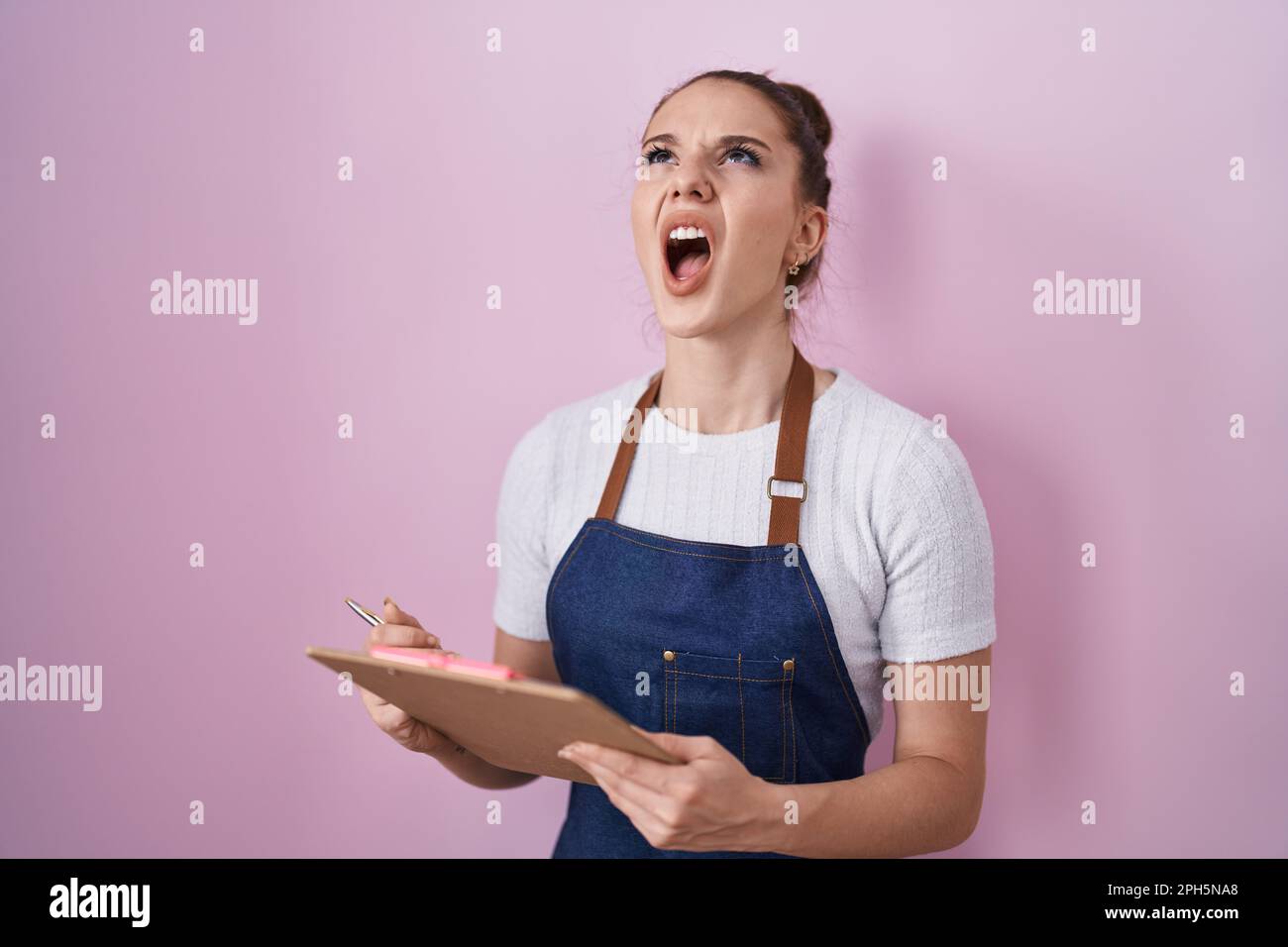 Young hispanic girl wearing professional waitress apron taking order angry and mad screaming ...