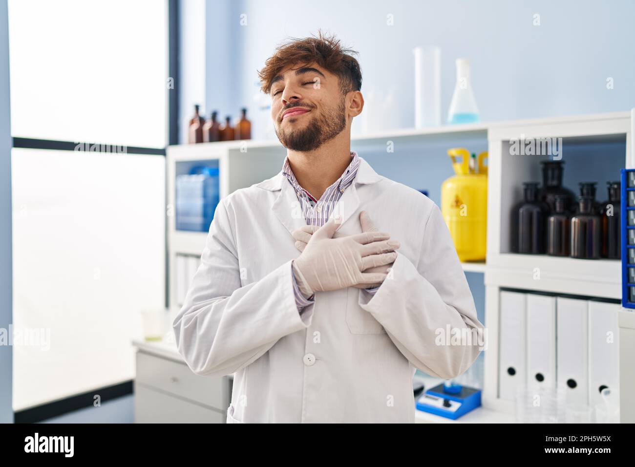 Arab man with beard working at scientist laboratory smiling with hands on chest, eyes closed ...