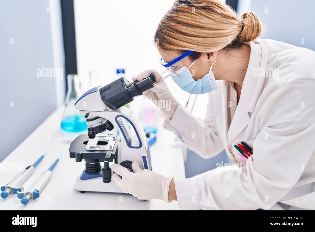 Young blonde woman scientist wearing medical mask using microscope at ...