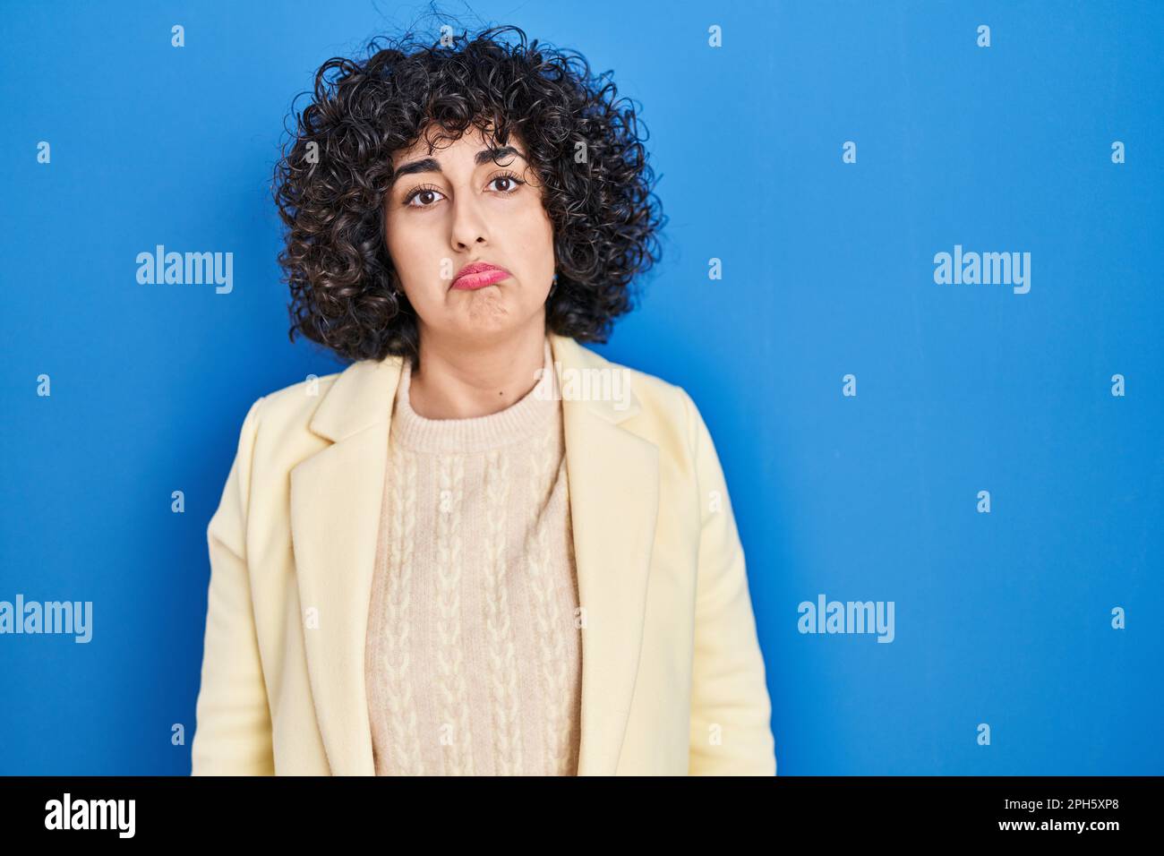 Young brunette woman with curly hair standing over blue background ...