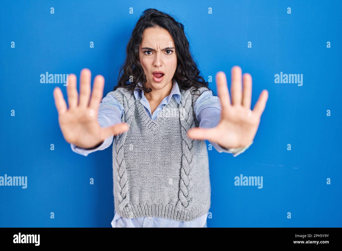 Young brunette woman standing over blue background doing stop gesture ...