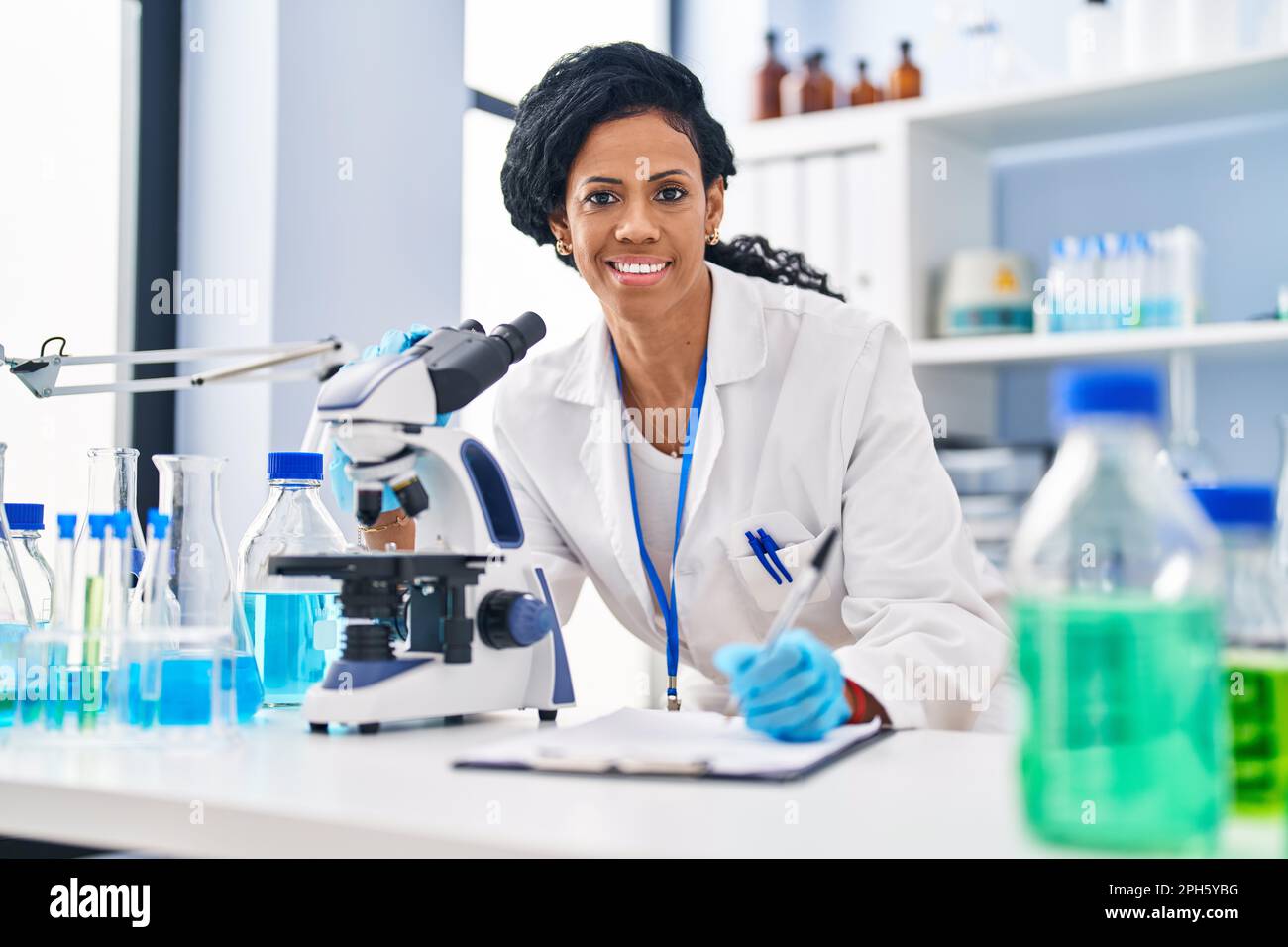African american woman wearing scientist uniform using microscope write on document at ...