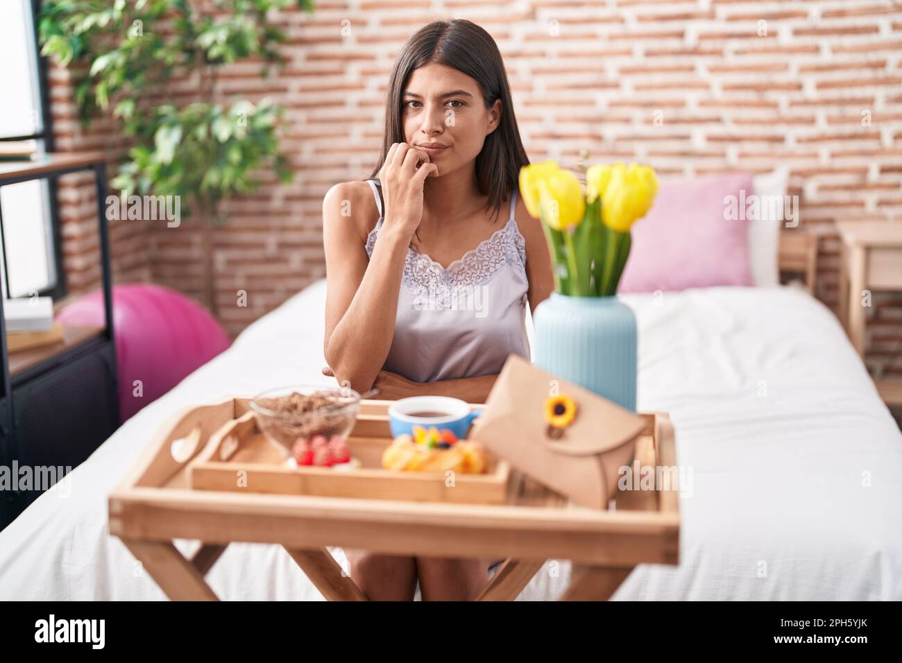 Brunette young woman eating breakfast sitting on the bed looking stressed and nervous with hands ...
