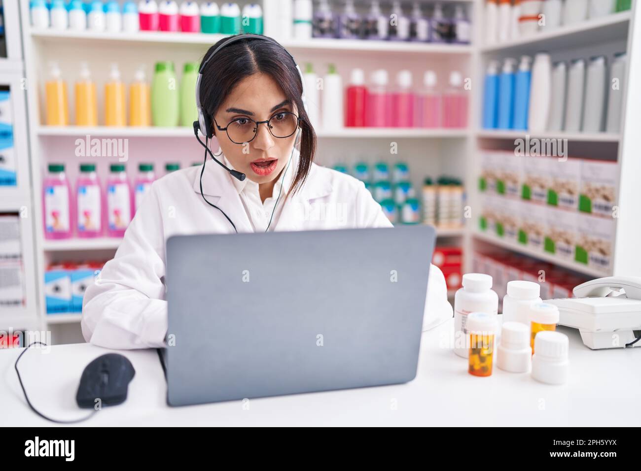 Young arab woman working at pharmacy drugstore using laptop afraid and shocked with surprise ...