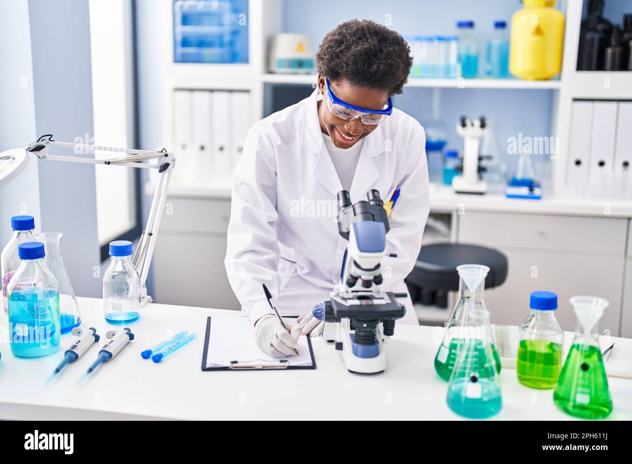 African american woman wearing scientist uniform using microscope write on clipboard at ...