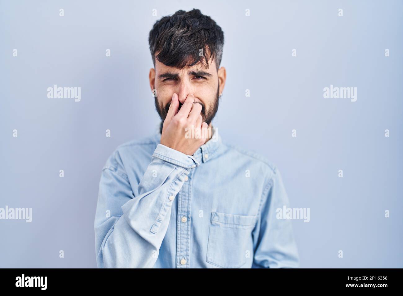 Young hispanic man with beard standing over blue background smelling ...