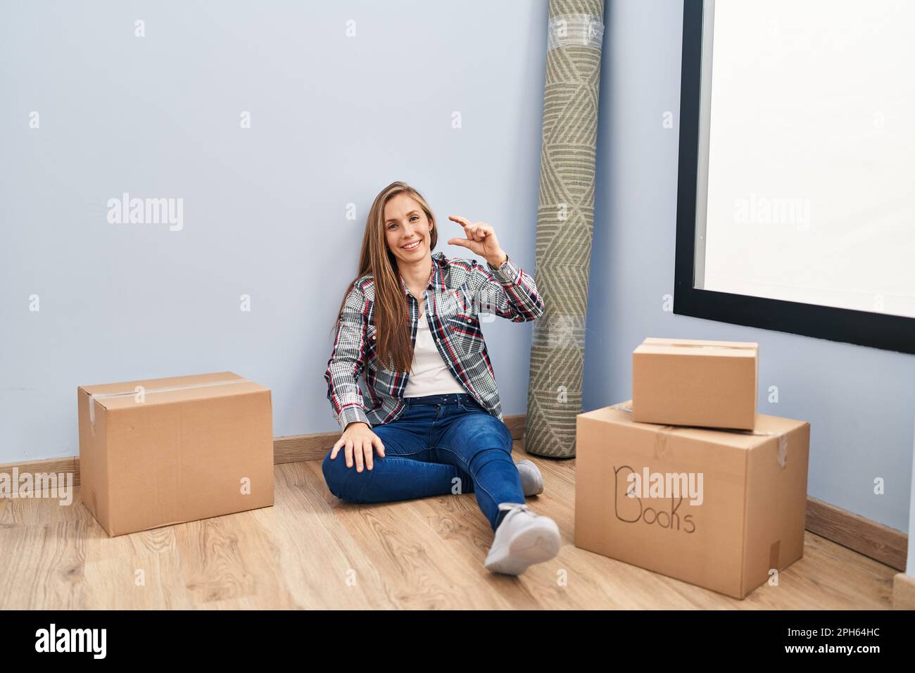 Young blonde woman sitting on the floor moving to a new home smiling ...