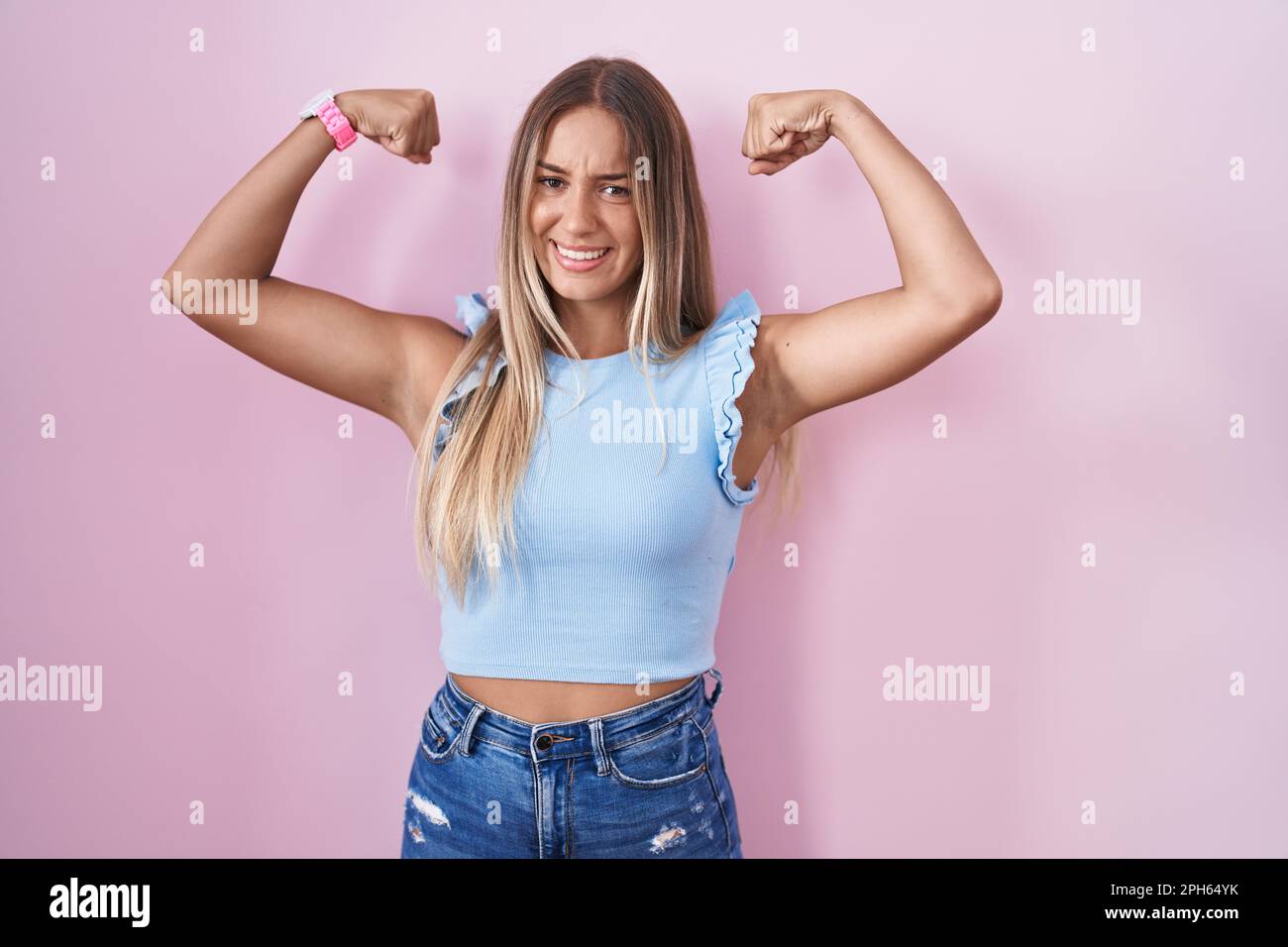 Young blonde woman standing over pink background showing arms muscles ...