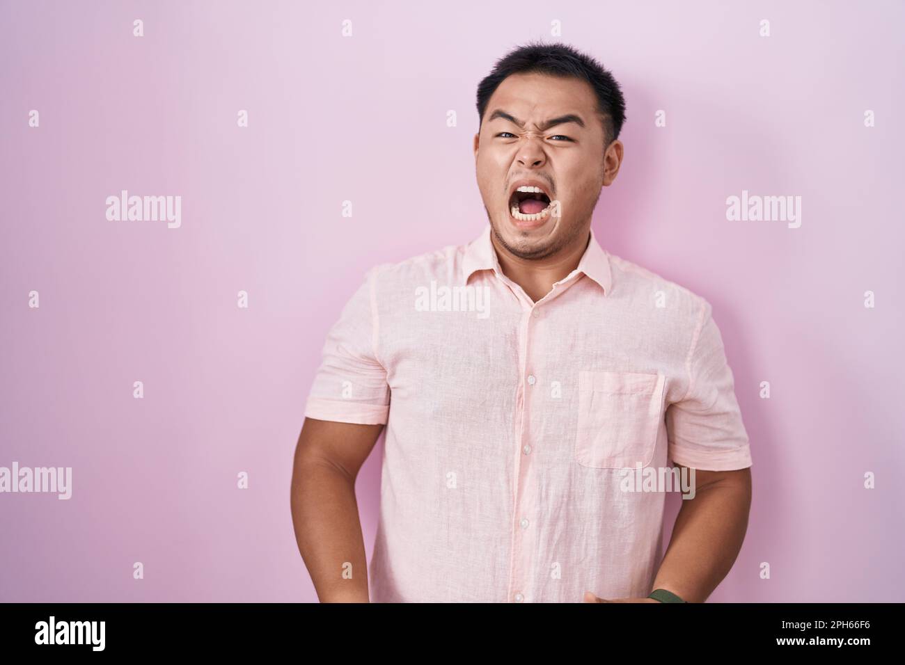 Chinese young man standing over pink background angry and mad screaming ...