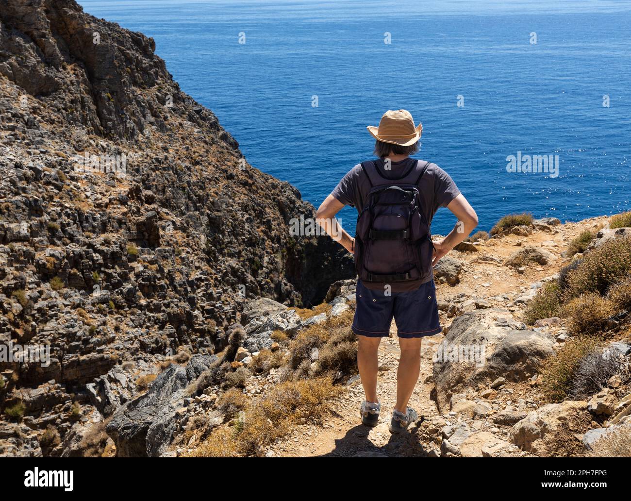 A long-haired man in a hat stands on the steep shore of a bright blue ...