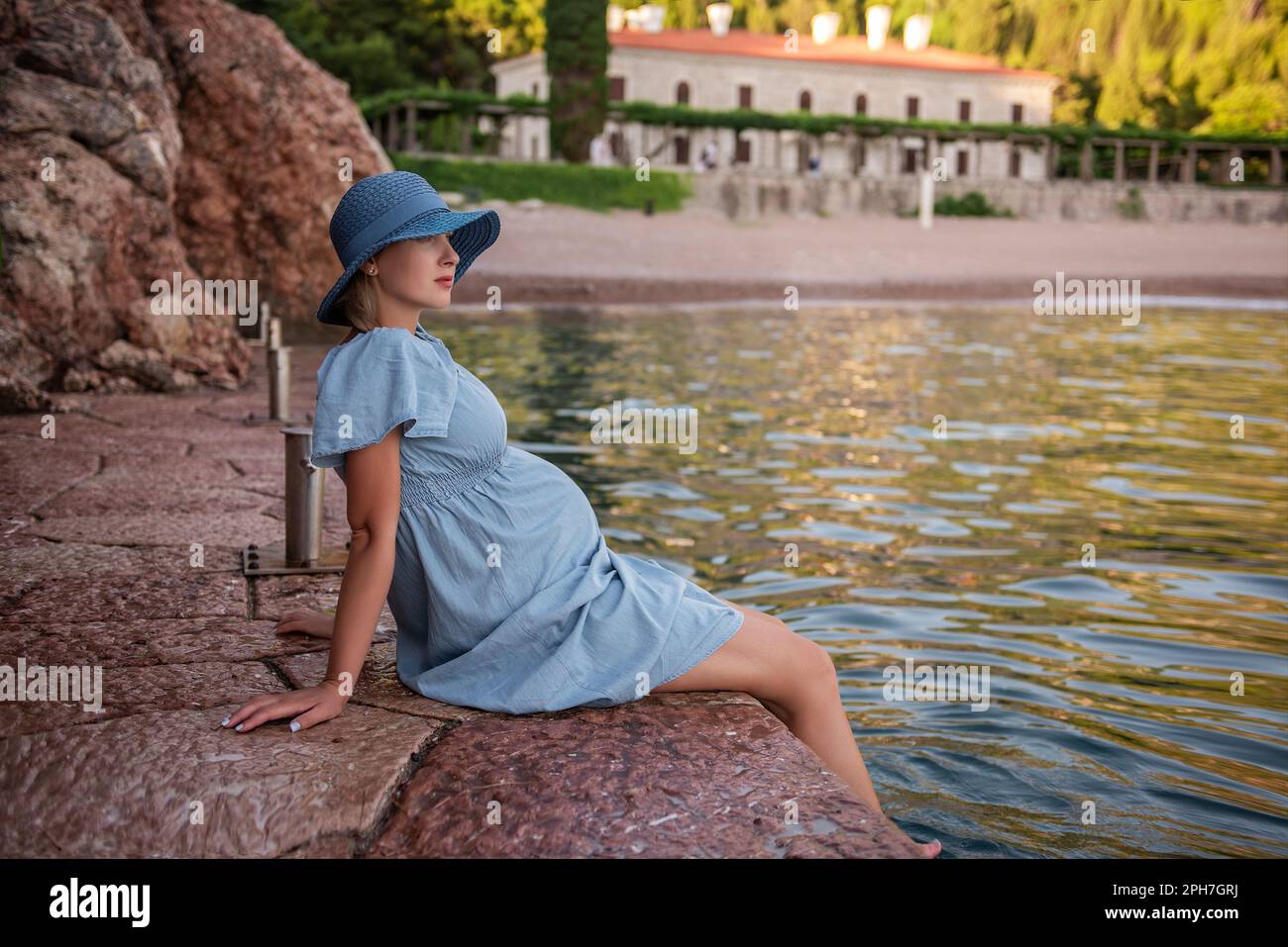 Pregnant woman traveler in denim dress, blue straw hat sits by the sea ...