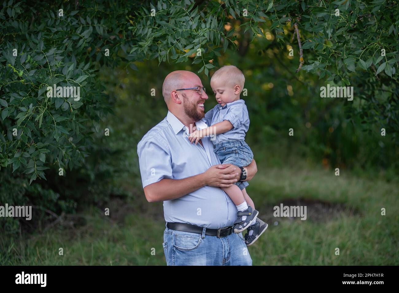 Bald man in glasses holds child in arms in the park. Father in jeans ...