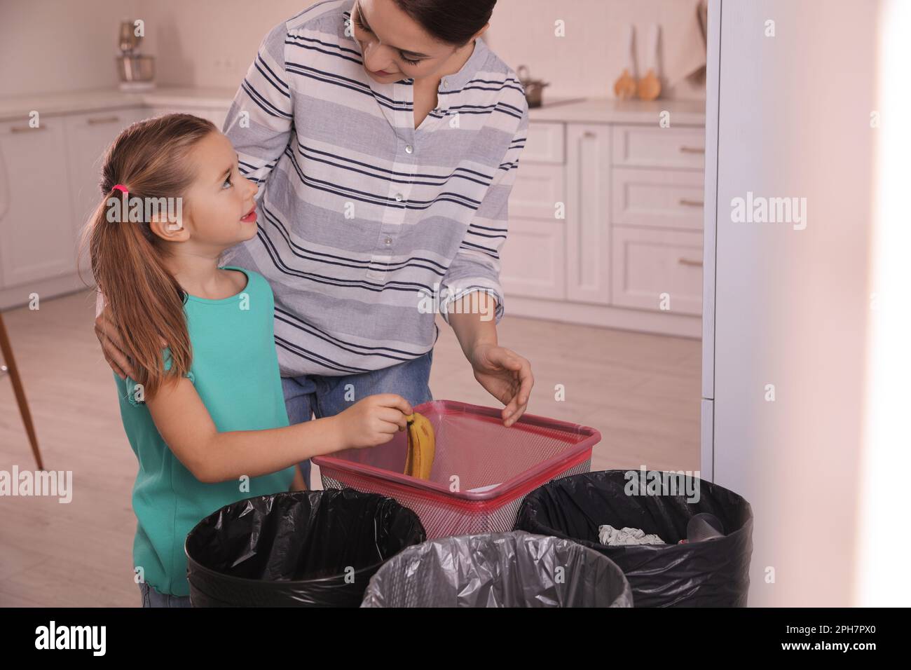 Young woman and her daughter throwing banana peel into trash bin in ...
