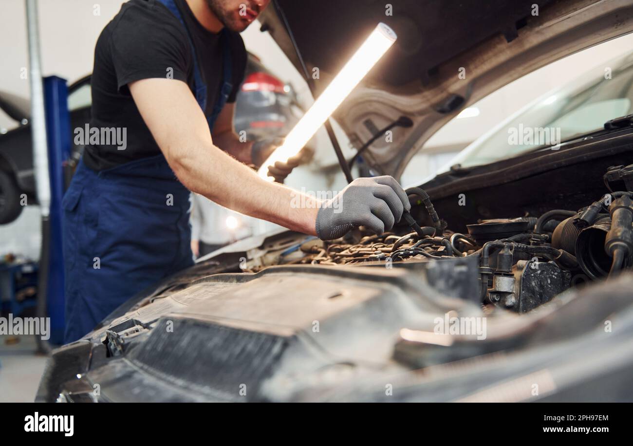 Uses lighting equipment. Man in work uniform repairs white automobile ...