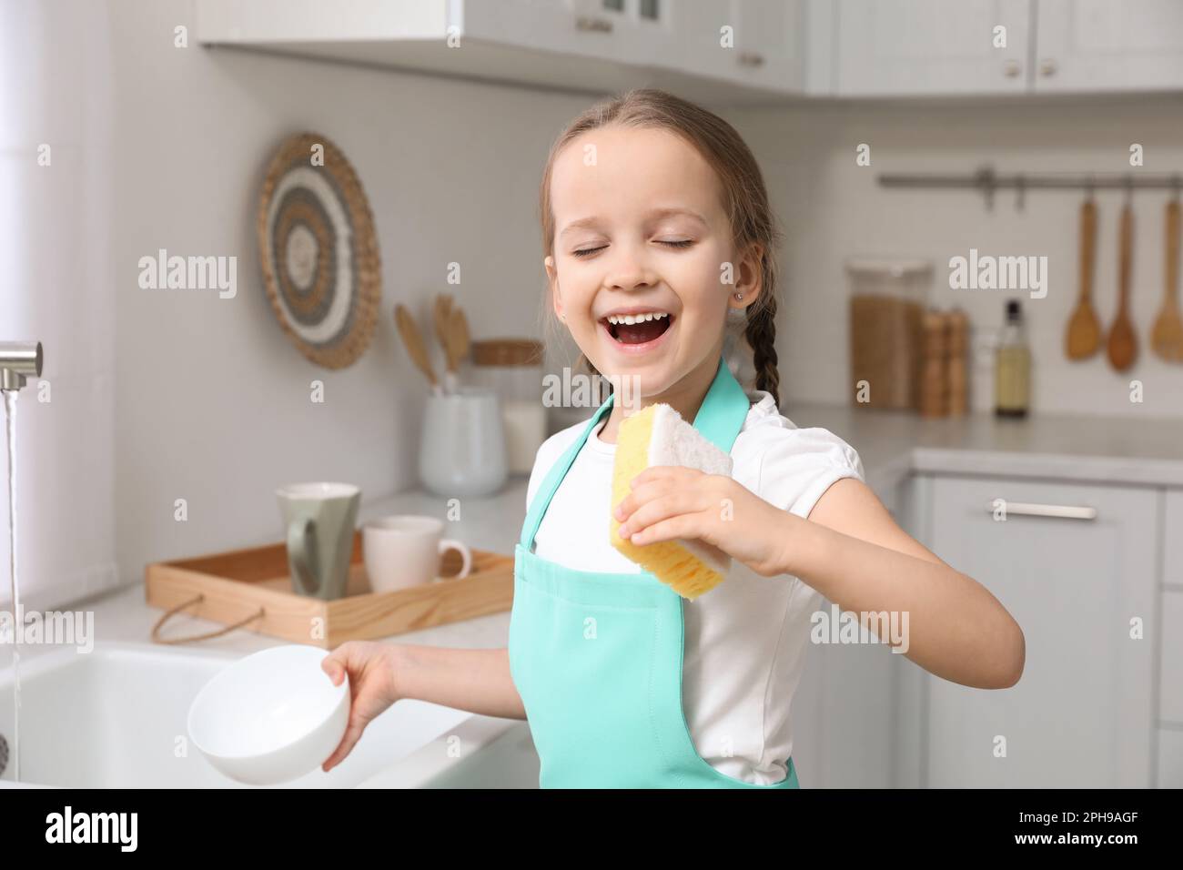 Cute little girl with sponge singing while washing dishes in kitchen ...