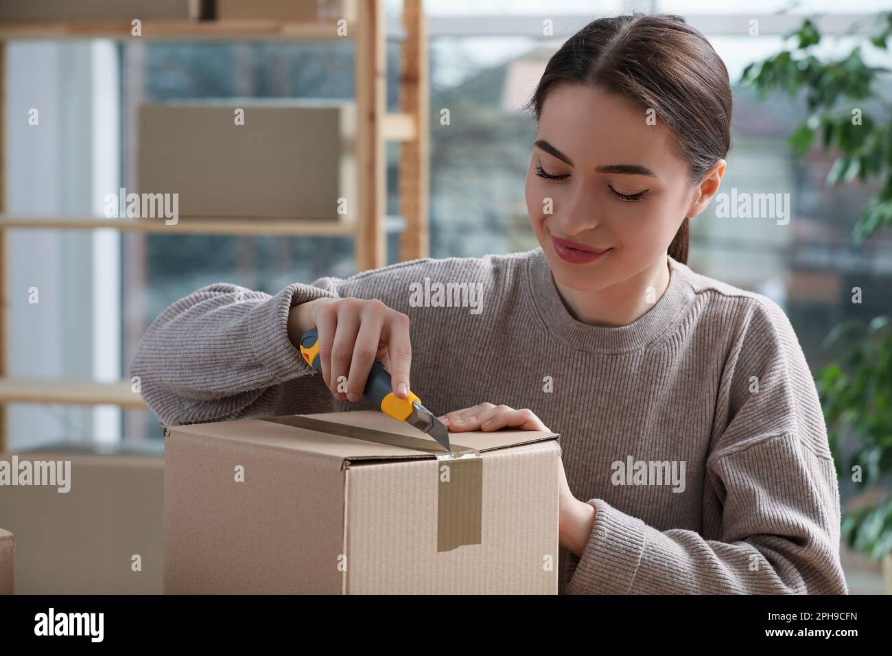 Young woman using utility knife to open parcel indoors Stock Photo - Alamy