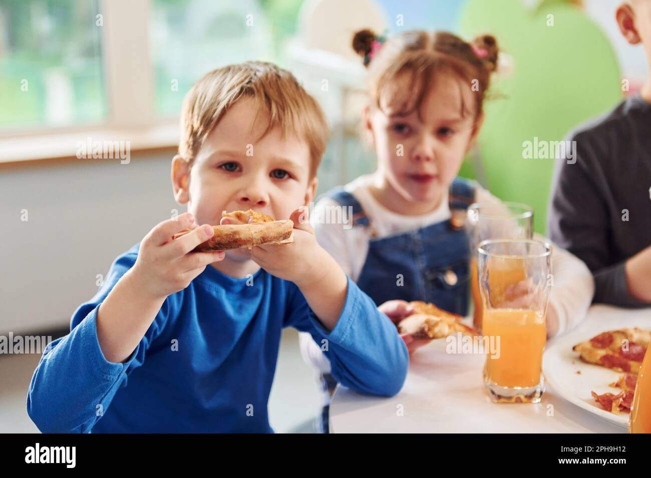 Three children sitting indoors by the table and eating pizza with ...