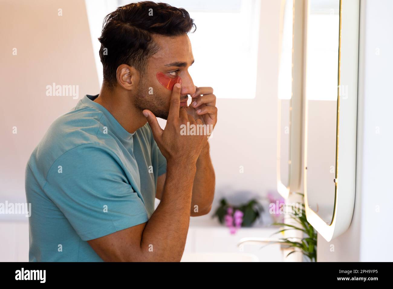 Biracial man looking in mirror and applying under eye mask in bathroom ...