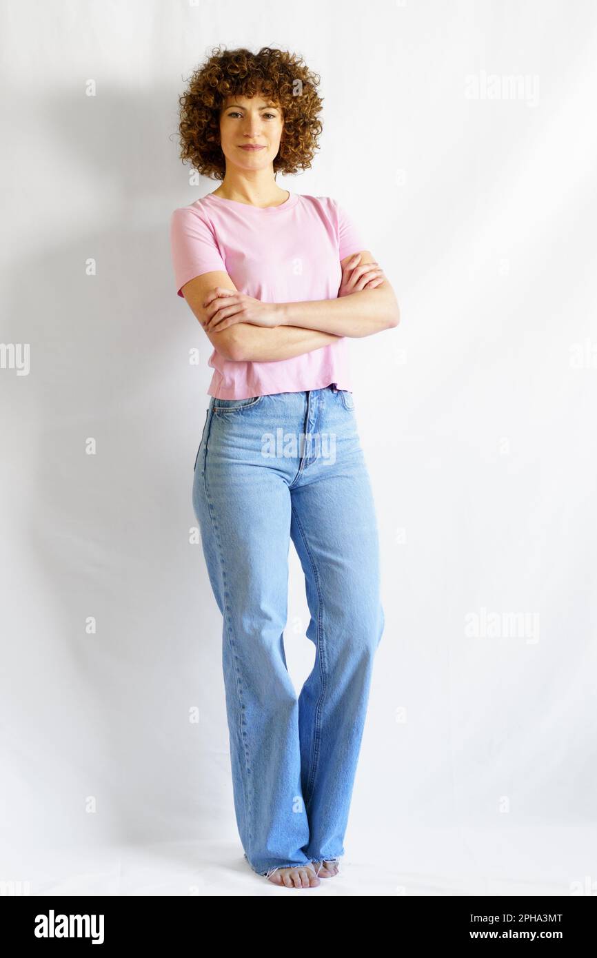 Full body of woman with curly hair standing against white wall crossing ...