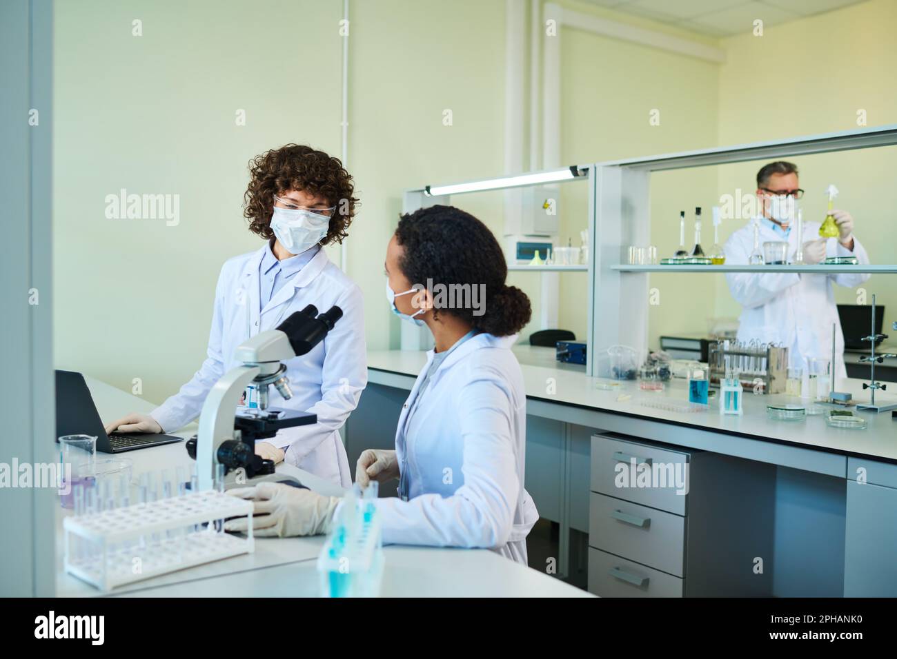 Two intercultural female virologists in lab coats and protective masks having discussion of ...