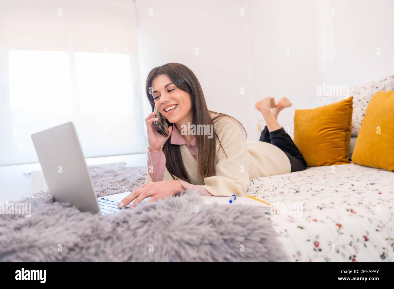 A female lying down on a bed while using her laptop and talking on her ...