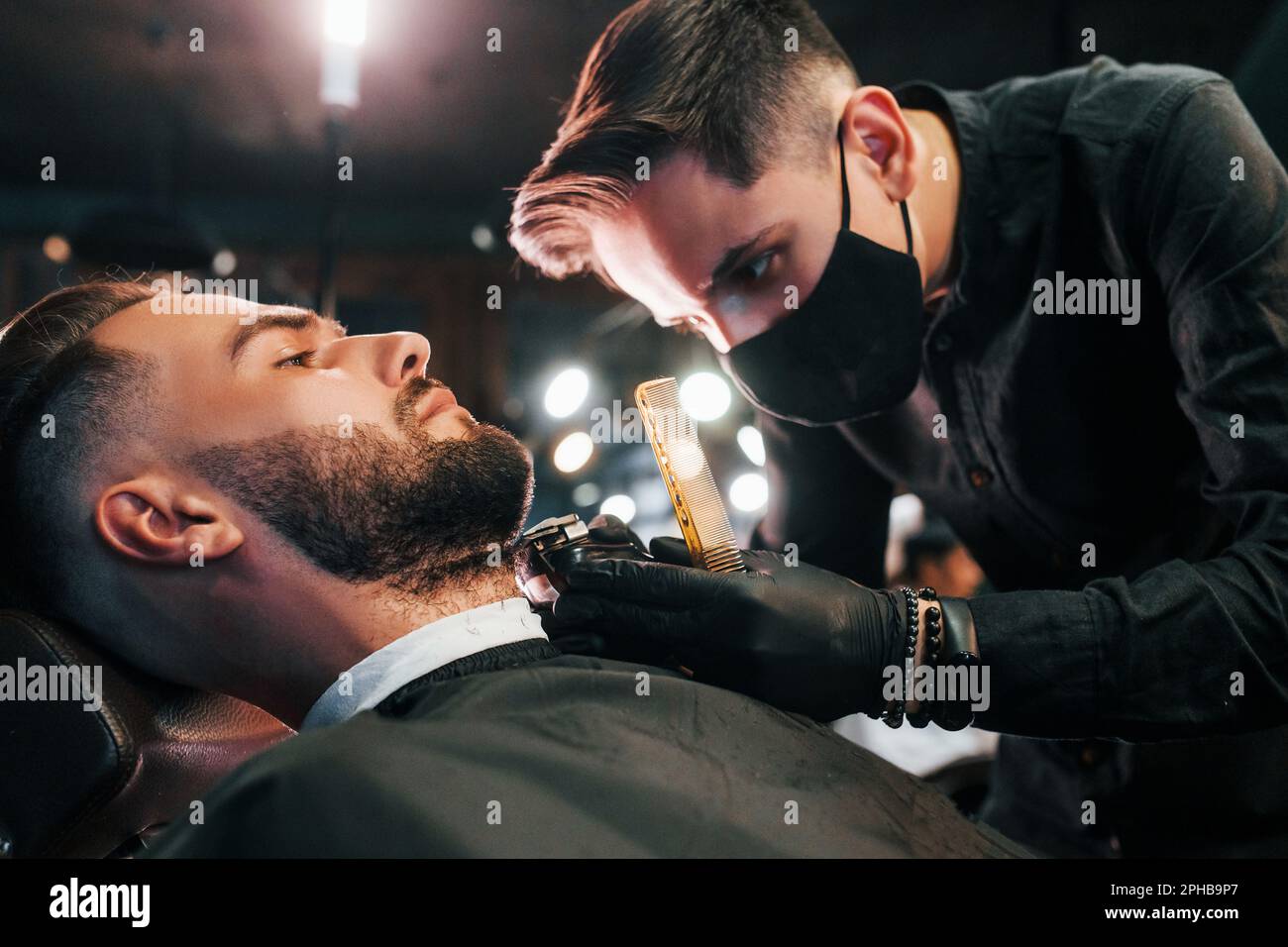 Young man with stylish hairstyle sitting and getting his beard shaved ...